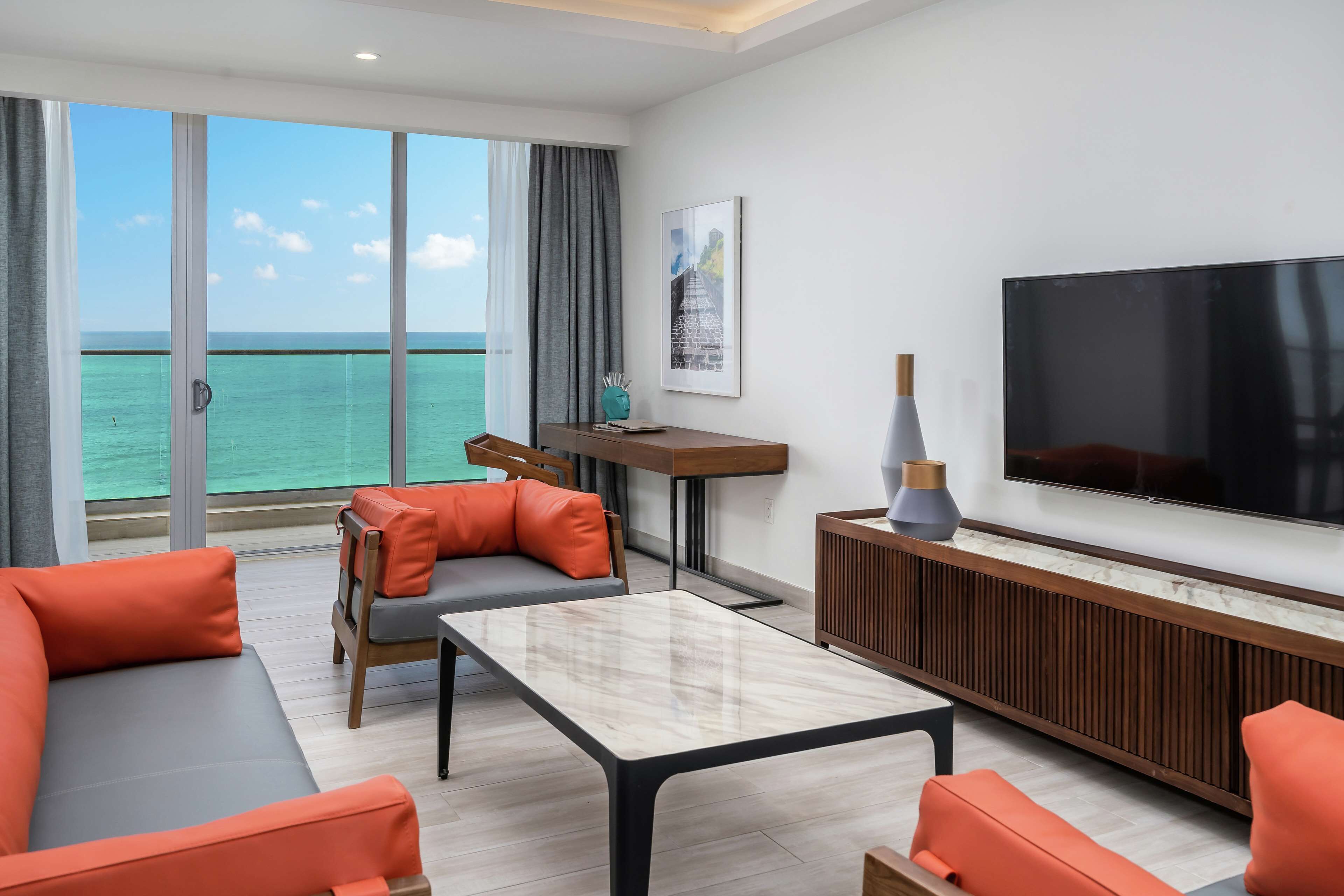 Uma sala de estar moderna com uma televisão grande, assentos confortáveis, uma mesa pequena e vista para o oceano através de amplas janelas.