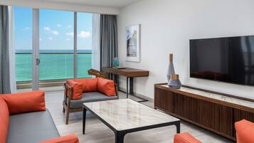 Uma sala de estar moderna com uma televisão grande, assentos confortáveis, uma mesa pequena e vista para o oceano através de amplas janelas.