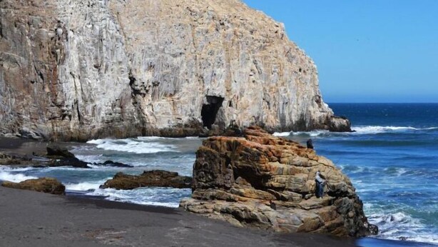 Litoral rochoso com um penhasco imponente e uma pessoa em uma formação rochosa.