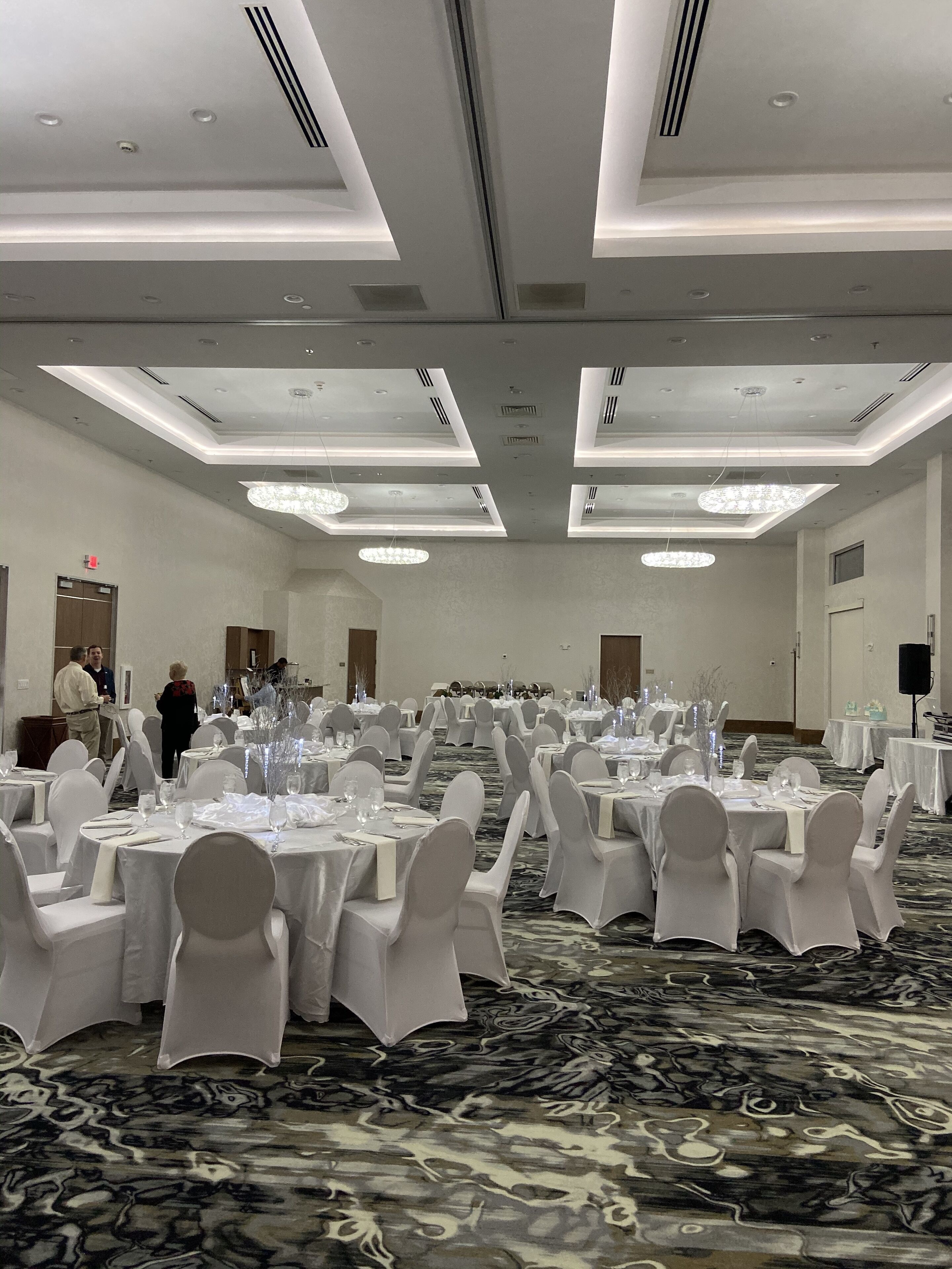 Uma sala de banquetes com mesas redondas, cadeiras brancas e um tapete estampado.