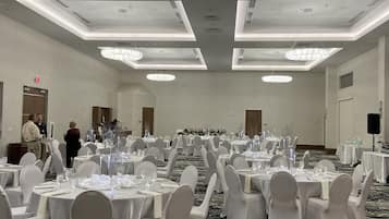 Uma sala de banquetes com mesas redondas, cadeiras brancas e um tapete estampado.