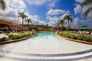 Uma piscina grande com borda em azulejo, cercada por espreguiçadeiras e palmeiras, sob um céu claro.