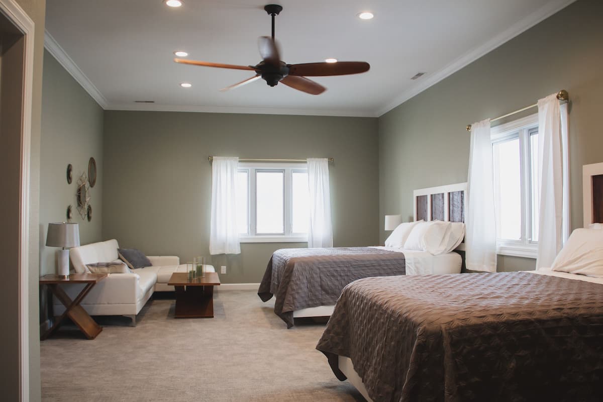 Quarto com duas camas, um ventilador de teto, uma janela com cortinas, uma mesa de cabeceira e um abajur.