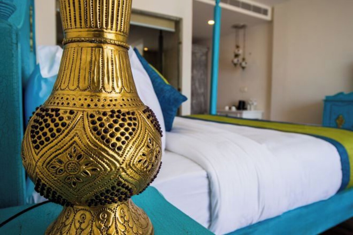 Uma cama com colcha branca e azul, um vaso decorativo dourado e móveis cor turquesa.