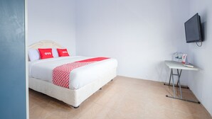 Uma cama bem arrumada com colcha branca e vermelha, uma pequena mesa de cabeceira com uma garrafa e uma caixa, e uma TV fixada na parede.