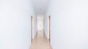 Um corredor comprido e estreito, com paredes brancas e chão de azulejos, que leva a uma porta no final.