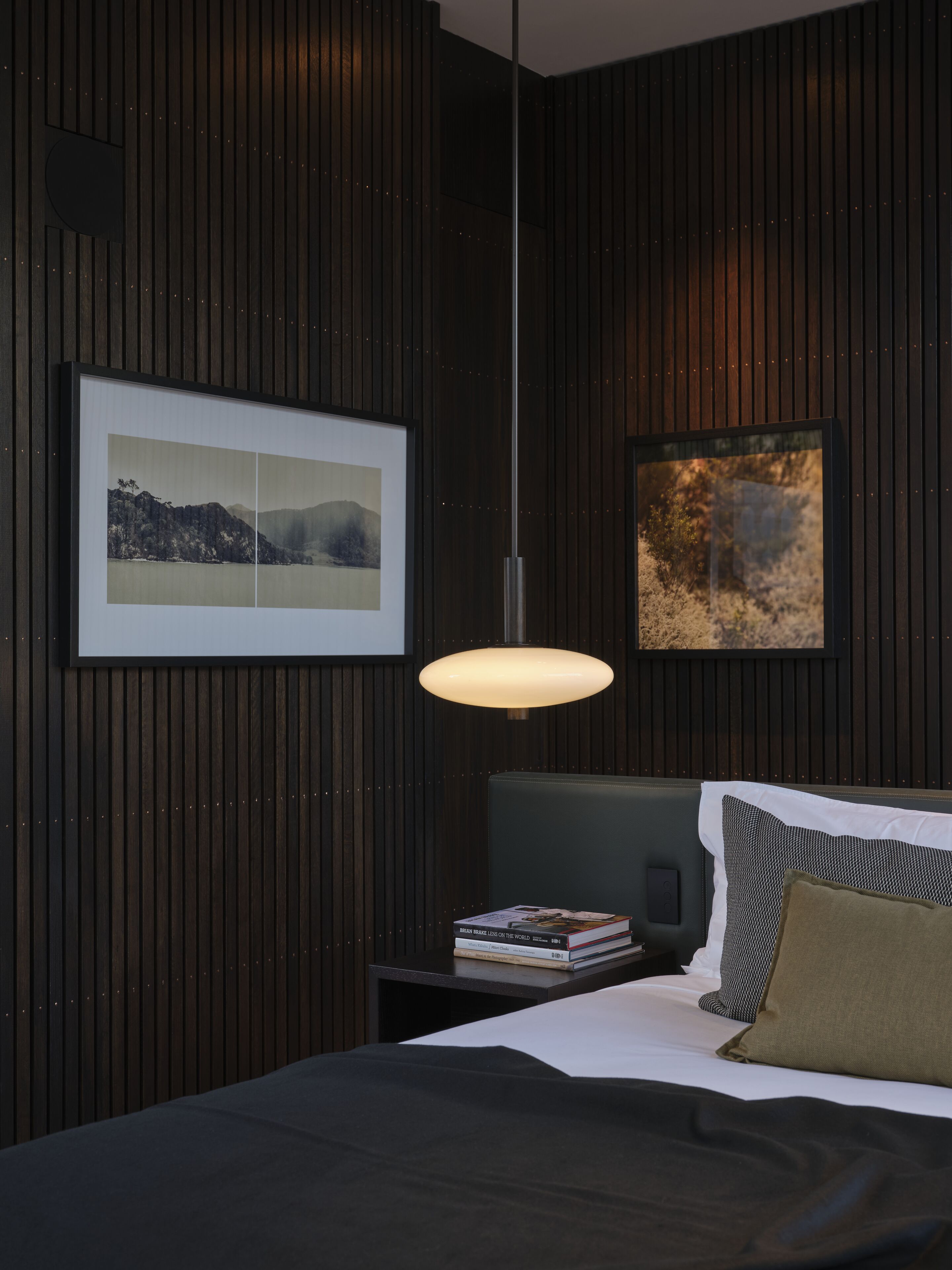 Quarto moderno com painéis de madeira escura, cama com cabeceira preta e branca, mesa de cabeceira com livros e duas fotografias emolduradas na parede.