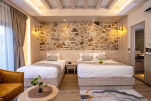 Quarto de hotel com duas camas, cabeceira de madeira, parede de pedra, uma pequena mesa com uma planta e um banheiro visível através de uma porta aberta.