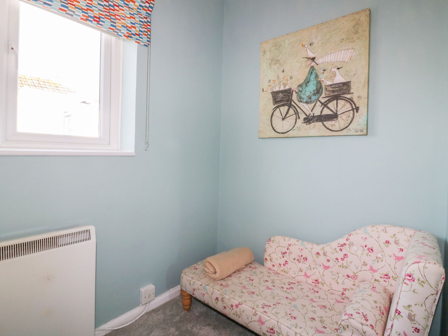 Um quarto com parede azul clara, janela com cortina estampada, radiador, sofá floral e um quadro de bicicleta.