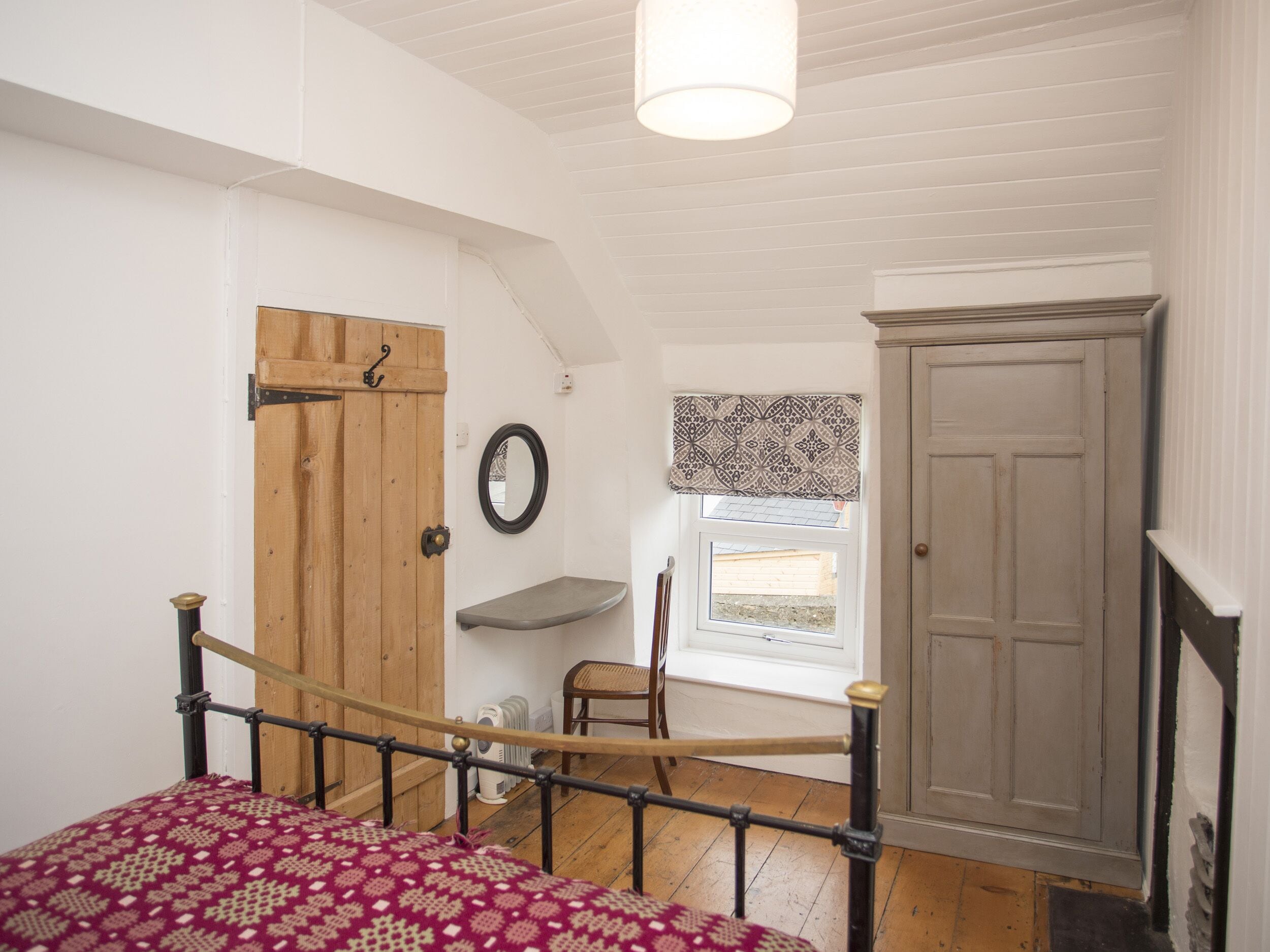 Quarto com uma cama, porta de madeira, janela com persiana estampada, radiador e uma cadeira.