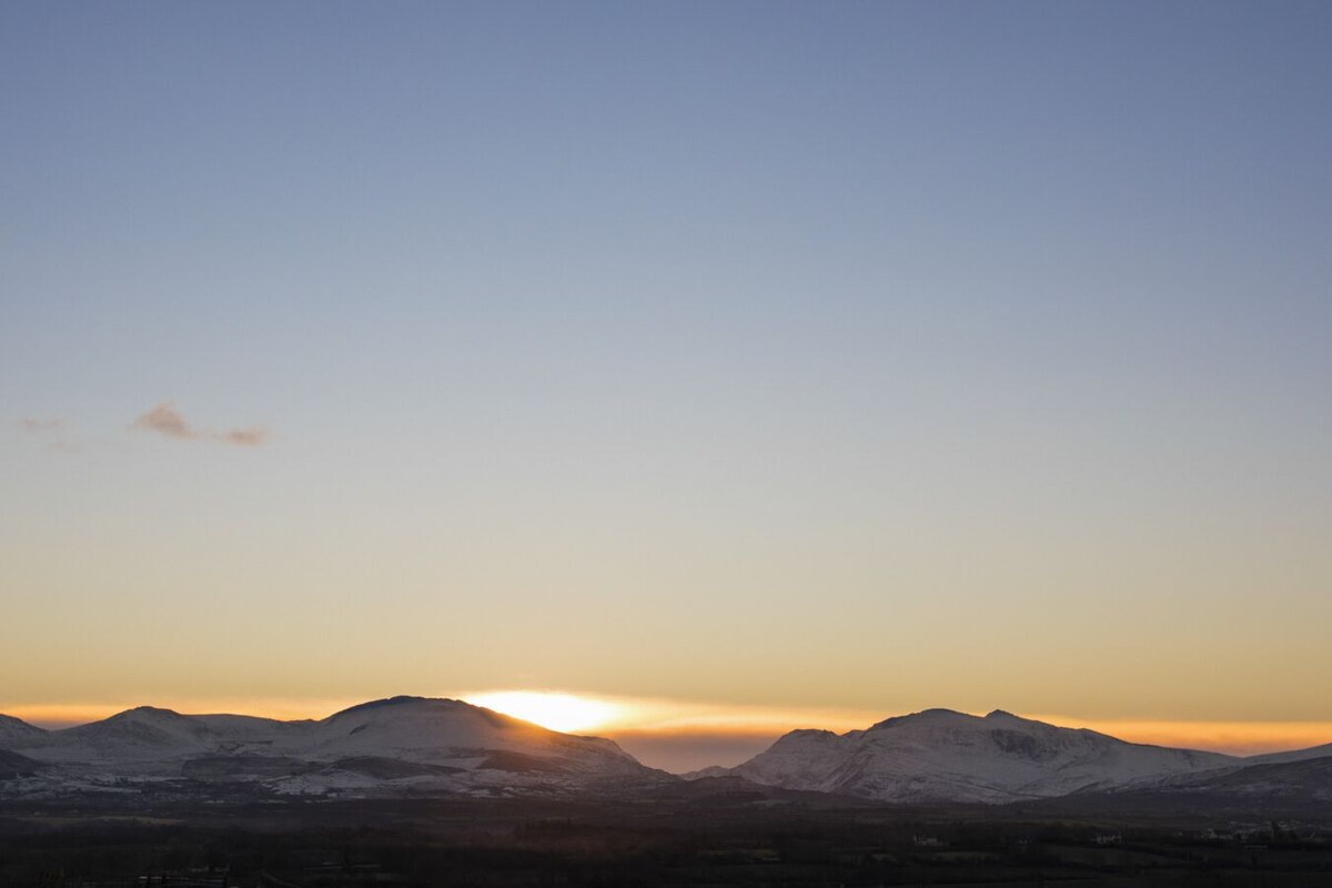 Paisagem montanhosa ao pôr do sol, com picos cobertos de neve e céu claro.