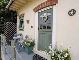 Uma casa charmosa com porta verde, um pequeno alpendre com uma cadeira azul e uma mesa, e um vaso com flores brancas.