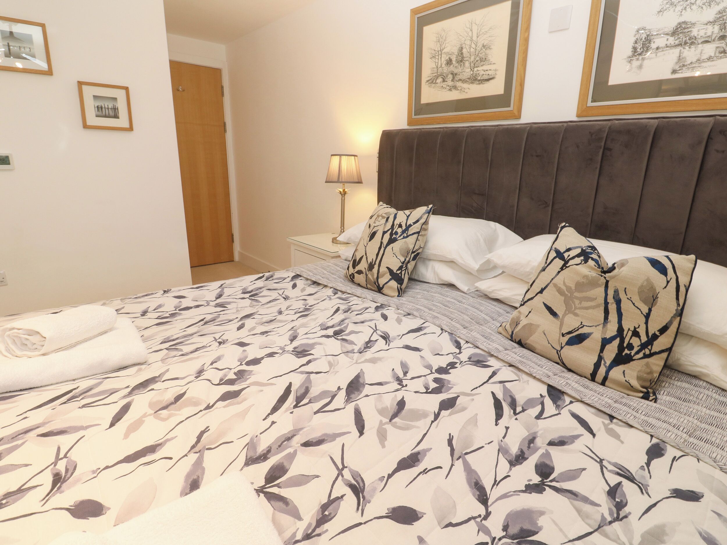 Quarto de hotel com cama, mesa de cabeceira, abajur e quadros nas paredes.