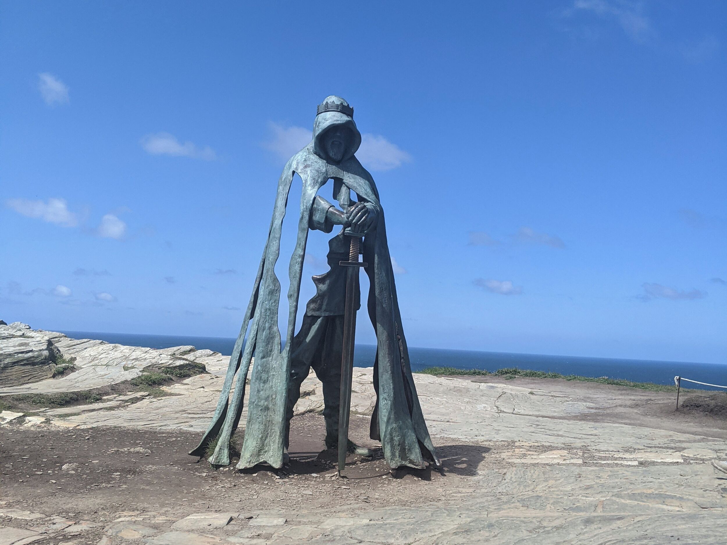 Uma estátua de uma figura com coroa e espada, sobre terreno rochoso, com céu claro e oceano ao fundo.