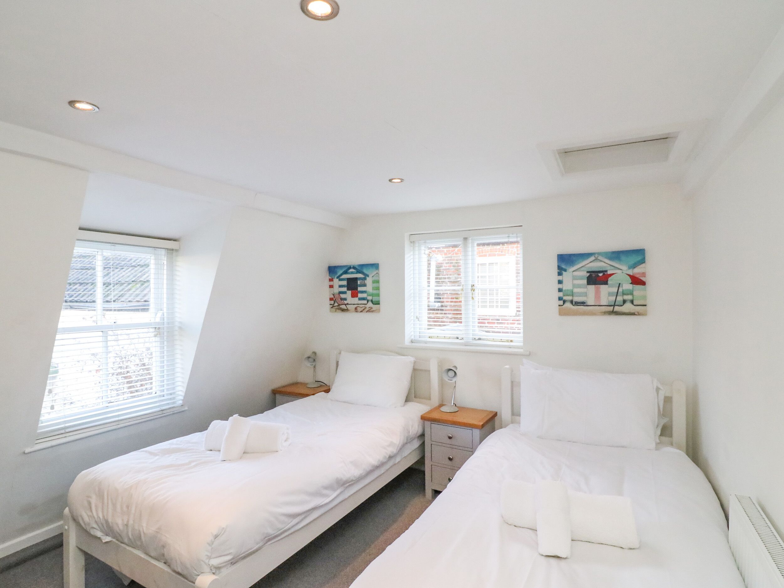 Quarto com duas camas de solteiro, janela com persianas, luminária no teto e duas pinturas penduradas na parede.