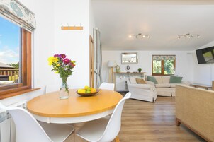 Uma sala de jantar com uma mesa de madeira, cadeiras brancas, um vaso de flores e uma tigela de frutas.