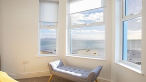 Quarto com vista para o mar através de grandes janelas, um banco acolchoado cinza e um cobertor amarelo no chão.