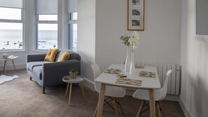 Uma sala de estar moderna com um sofá cinzento, uma mesinha redonda com uma planta, e uma área de jantar com uma mesa e cadeiras brancas.