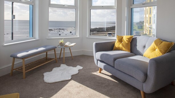 Sala moderna com um sofá cinzento, um banquinho e uma mesa pequena com um livro e uma planta. Grandes janelas oferecem vista para a praia e o mar.