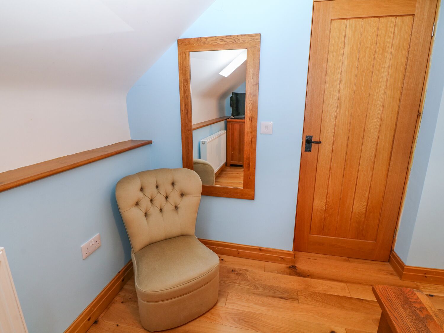Um quarto com piso de madeira, uma cadeira estofada bege e uma porta de madeira, além de um espelho na parede.