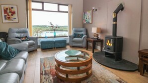 Uma sala de estar com sofá, duas poltronas, uma mesa de centro de vidro, lareira e uma janela grande com vista.