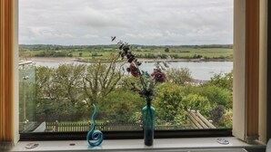 Uma janela com vista para um rio e vegetação, um vaso azul-esverdeado com flores, e uma escultura de vidro azul num parapeito.