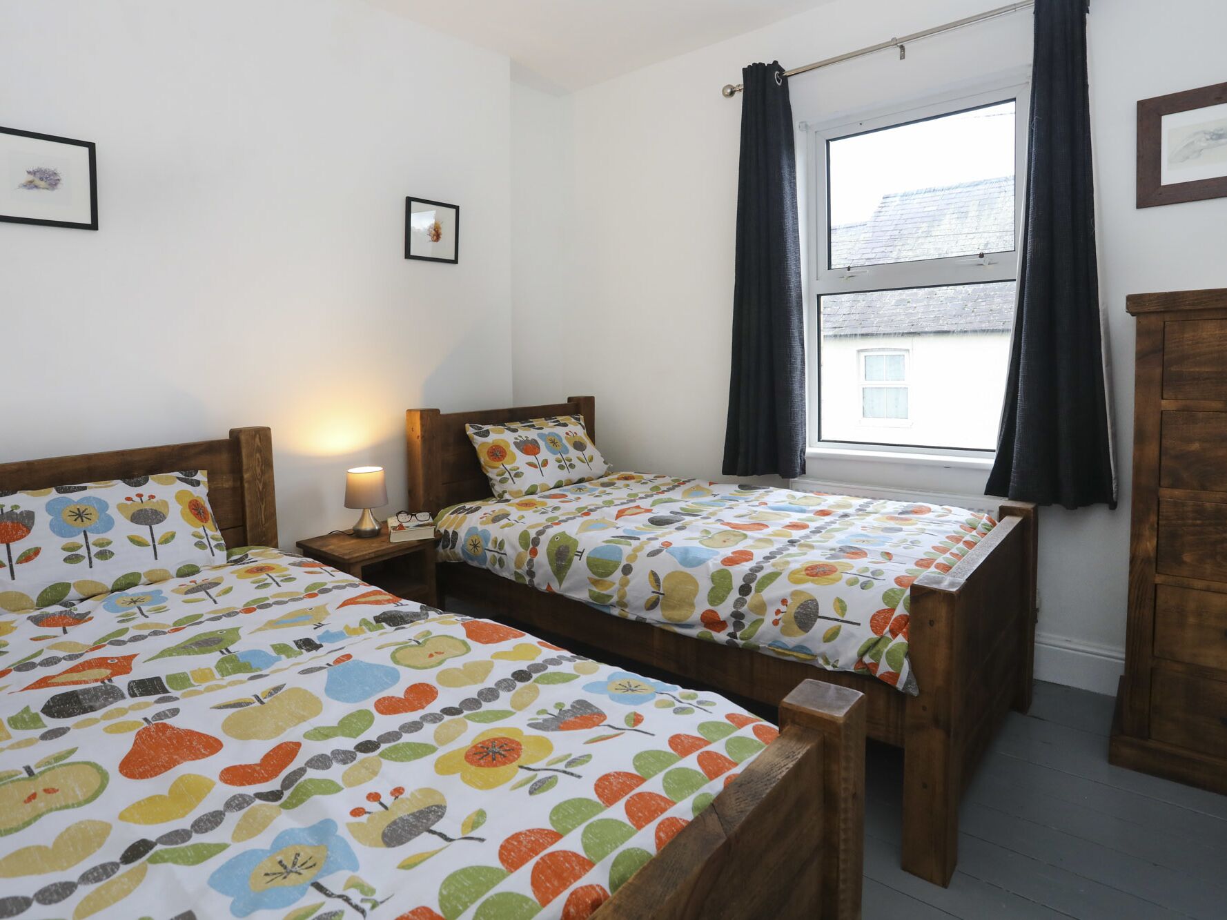 Quarto com duas camas de madeira, uma janela com cortinas e uma mesa de cabeceira com abajur.