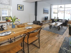 Uma sala de jantar com uma mesa de madeira posta para quatro pessoas, rodeada por cadeiras. Há uma televisão sobre um suporte à direita, e um sofá ao fundo.