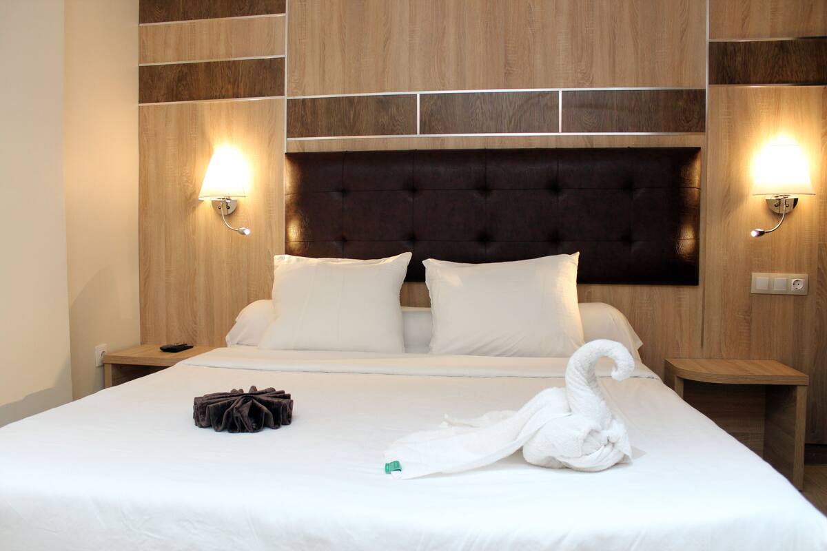 Uma cama bem arrumada com lençóis e almofadas brancas, uma cabeceira escura e duas mesas de cabeceira com abajures.