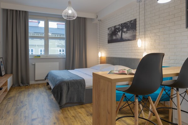Quarto com cama, escrivaninha de madeira, duas cadeiras, janela com cortinas, luminária na parede e um quadro.