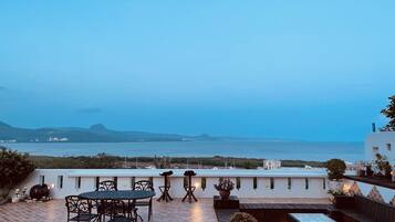 Um terraço com mesa e cadeiras, plantas em vasos e vista para o mar e colinas ao longe.