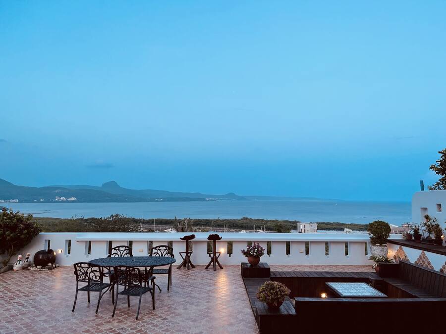 Um terraço com mesa e cadeiras, plantas em vasos e vista para o mar e colinas ao longe.