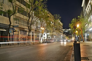 Cena noturna de rua com edifícios, árvores e postes de luz.