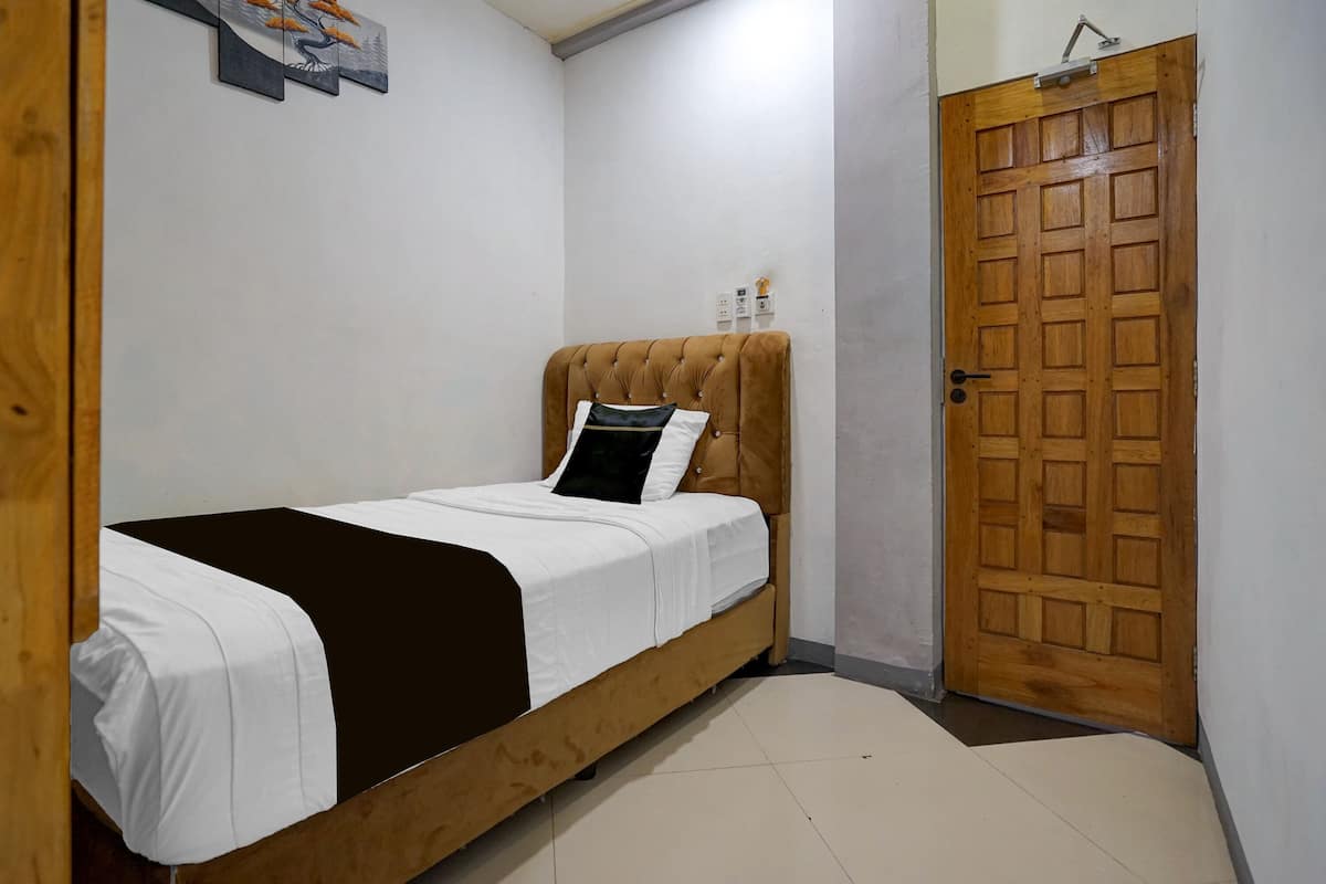 Uma cama de solteiro com cabeceira de madeira, colchão branco e manta preta. Uma porta de madeira com padrão de grade à direita.