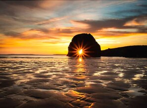 Um pôr do sol sobre uma costa rochosa com uma formação rochosa em destaque no centro.
