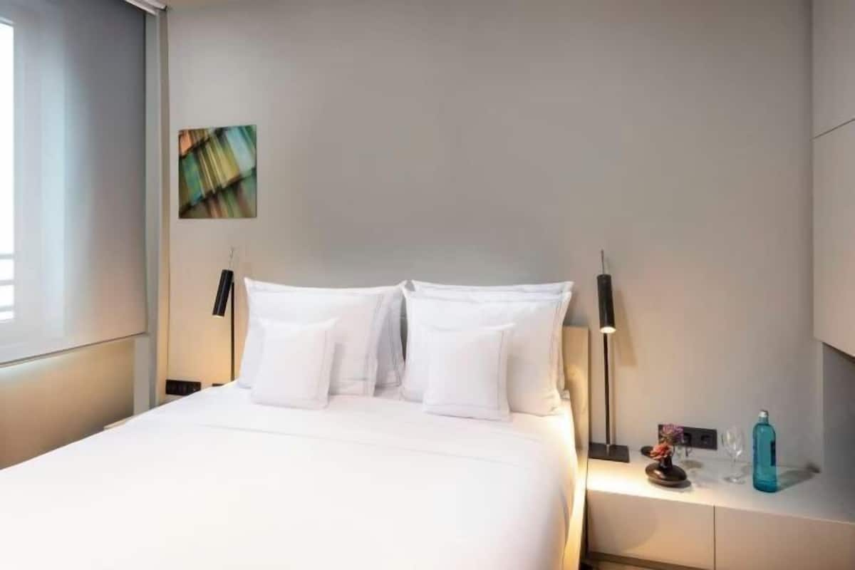 Quarto de hotel com cama, roupa de cama branca, mesa de cabeceira com abajur, pequena mesa com garrafa e copos, e uma obra de arte na parede.