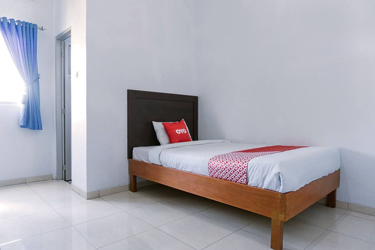 Uma cama de solteiro com estrutura de madeira, roupa de cama branca e almofada vermelha com a inscrição 'oyo'.