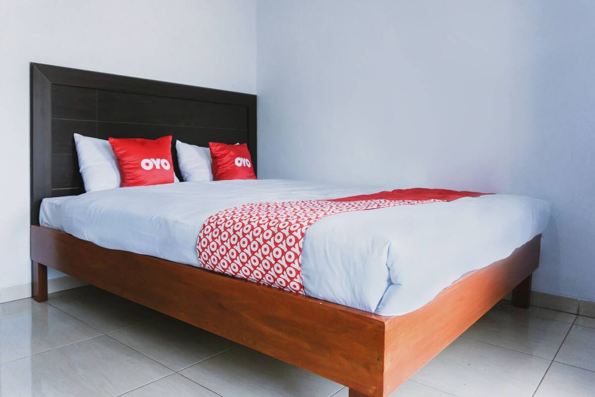 Quarto de hotel moderno com cama de madeira, roupa de cama branca e almofadas vermelhas com um logotipo.