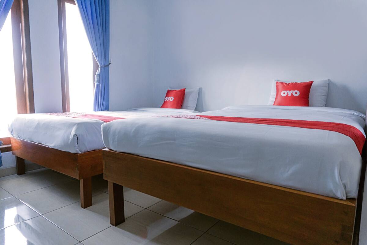 Duas camas de casal com roupa de cama branca e vermelha, uma cabeceira de madeira e uma cortina azul.