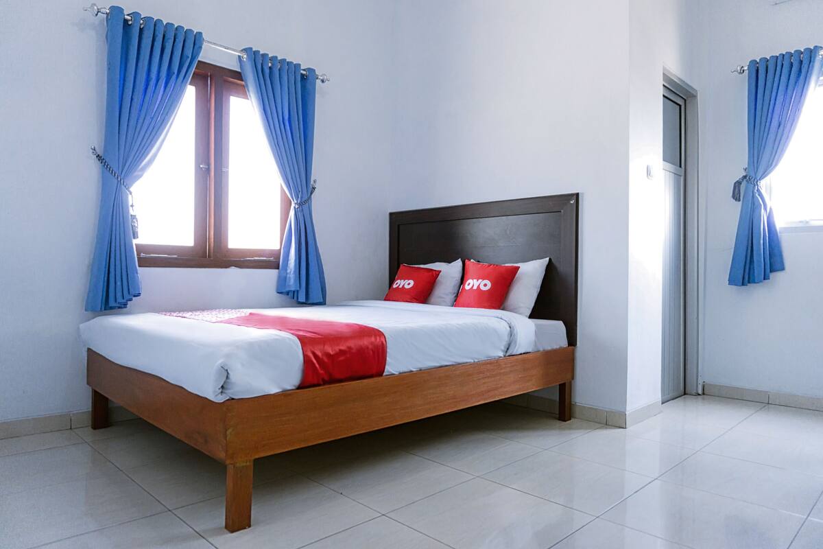 Uma cama de solteiro com estrutura de madeira, roupa de cama branca e almofadas vermelhas e brancas.