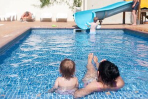 Piscina com uma criança e um adulto na água, um escorregador e cadeiras de descanso ao fundo.