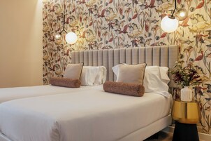 Uma cama de casal com roupa de cama branca, duas almofadas decorativas, uma cabeceira estampada em xadrez de dentes de cão e um vaso com flores secas em uma mesa de cabeceira.