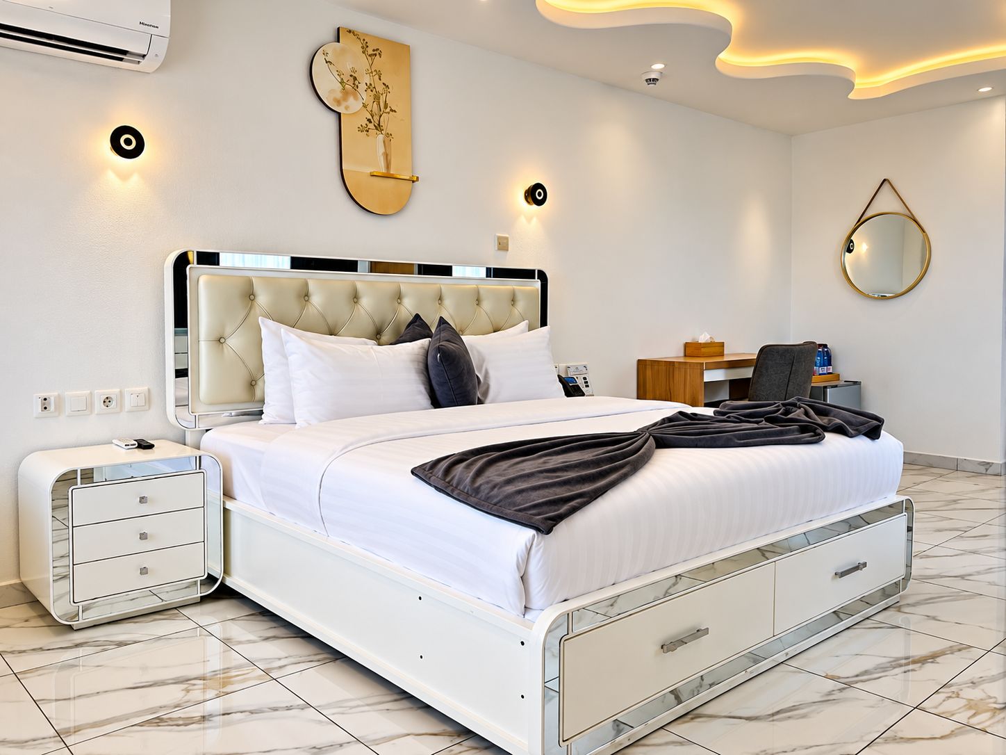 Quarto de hotel moderno com cabeceira estofada, mesa de cabeceira com gavetas, espelho e abajur.