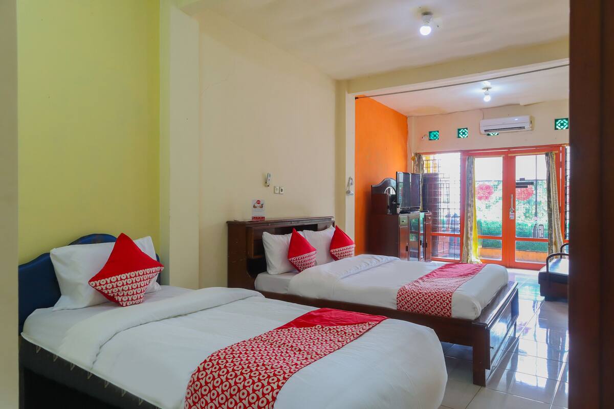 Quarto de hotel com duas camas, cada uma com roupa de cama branca e detalhes vermelhos, cabeceira de madeira e vista para o exterior por uma porta de vidro.