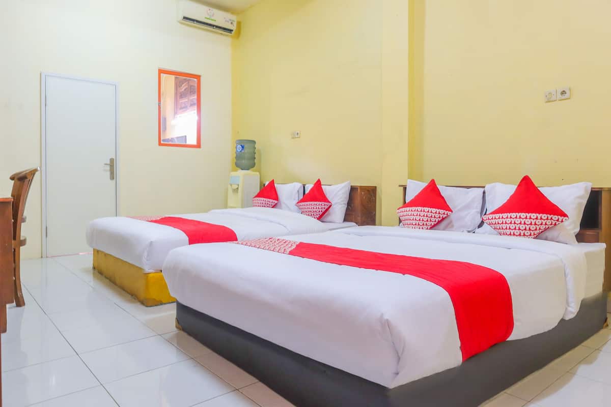 Quarto de hotel com duas camas, cada uma com roupa de cama branca e almofadas vermelhas, um dispensador de água e uma porta.