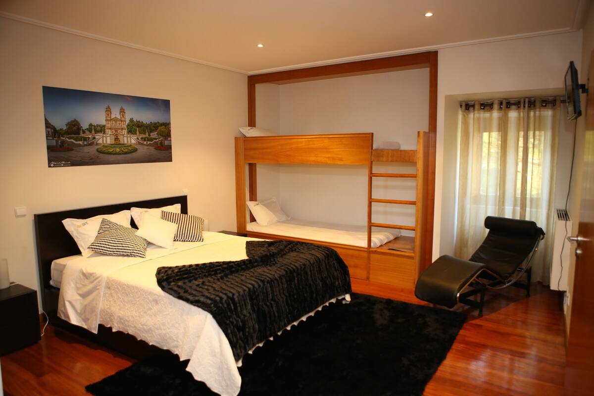 Quarto com cama beliche, uma cama de solteiro, uma poltrona de couro preto, tapete preto e uma pintura de um edifício na parede.
