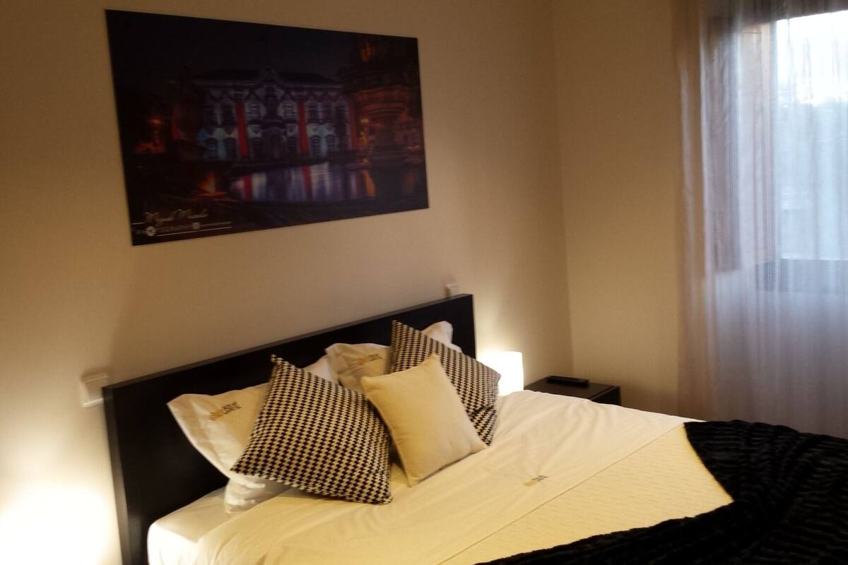 Quarto de hotel com uma cama, mesa de cabeceira, abajur e um quadro de um edifício na parede.