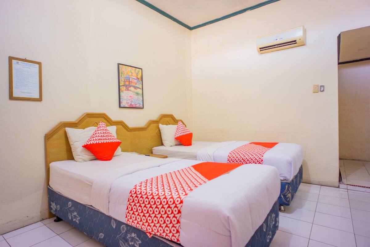 Quarto de hotel com duas camas, cada uma com colcha e almofadas estampadas. Há um certificado emoldurado na parede e um ar condicionado acima das camas.