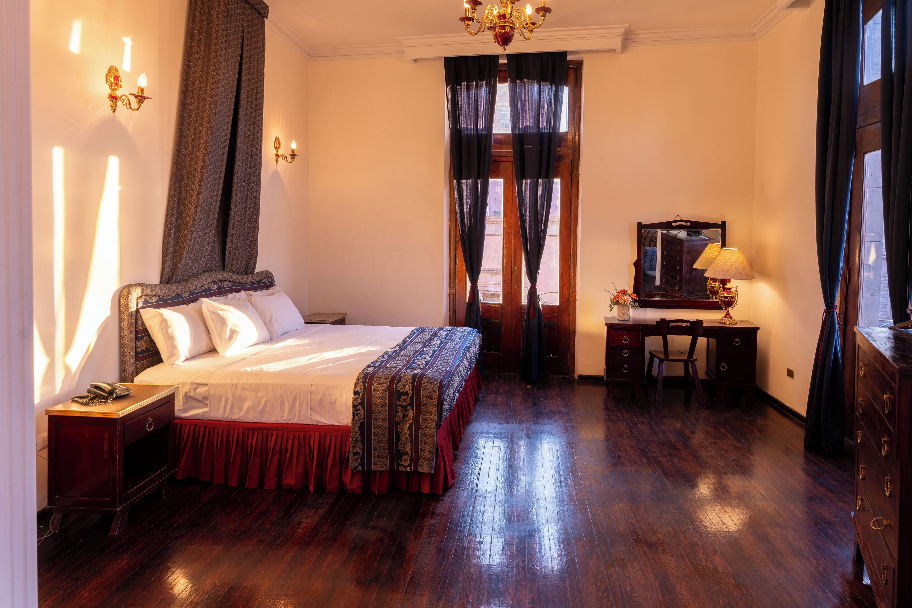 Quarto de hotel com uma cama grande, uma escrivaninha com luminária, um armário e um espelho.
