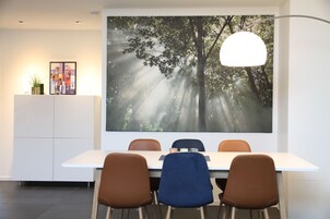 Uma zona de refeições moderna com uma grande obra de arte pendurada na parede retratando uma cena de floresta, uma mesa branca e cadeiras castanhas.